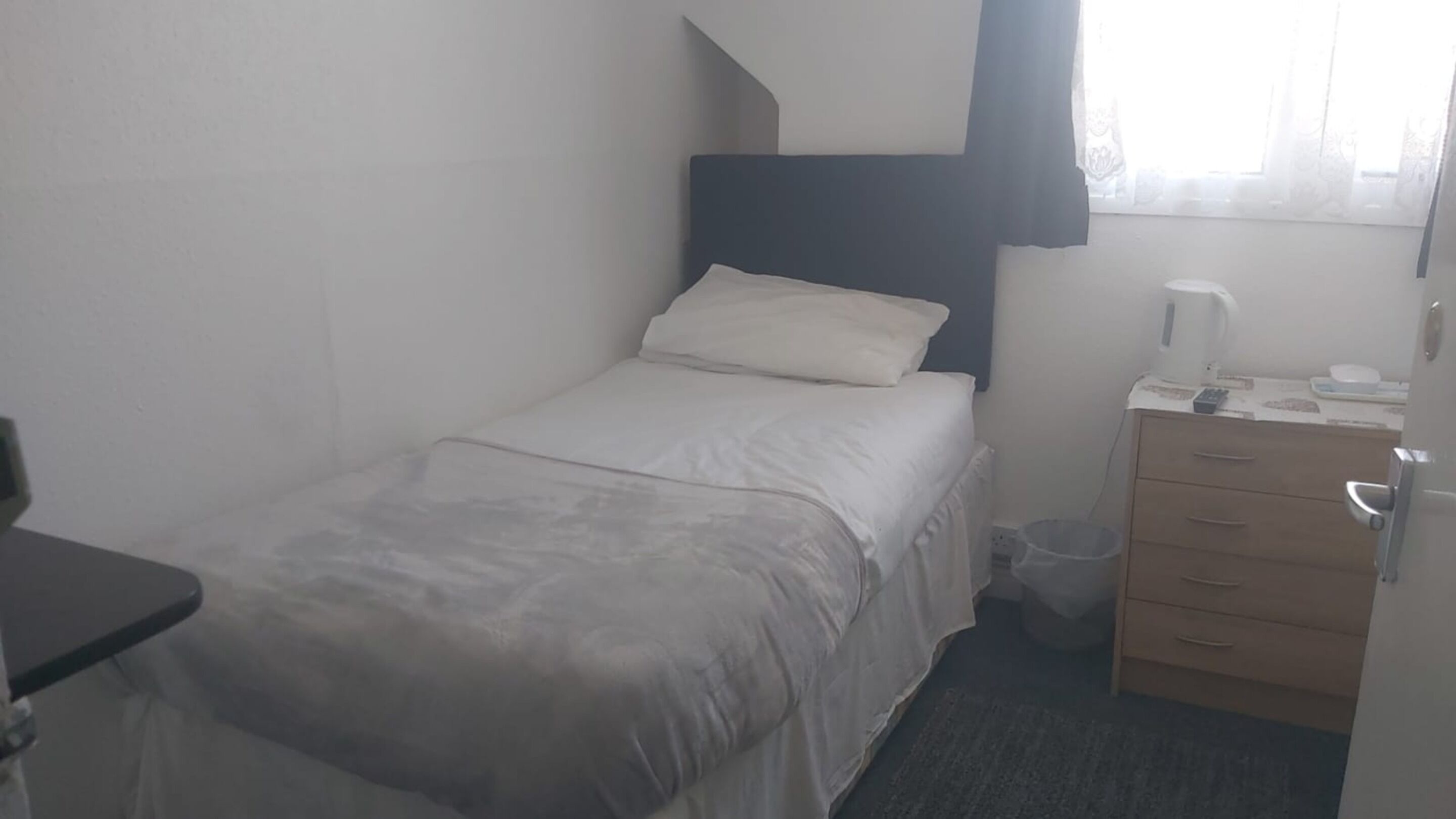 Quarto pequeno e bem organizado, com uma cama de solteiro, mesa de cabeceira com abajur, cômoda e uma janela com cortinas leves.