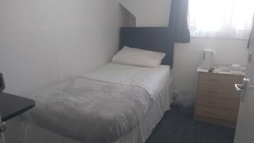 Quarto pequeno e bem organizado, com uma cama de solteiro, mesa de cabeceira com abajur, cômoda e uma janela com cortinas leves.