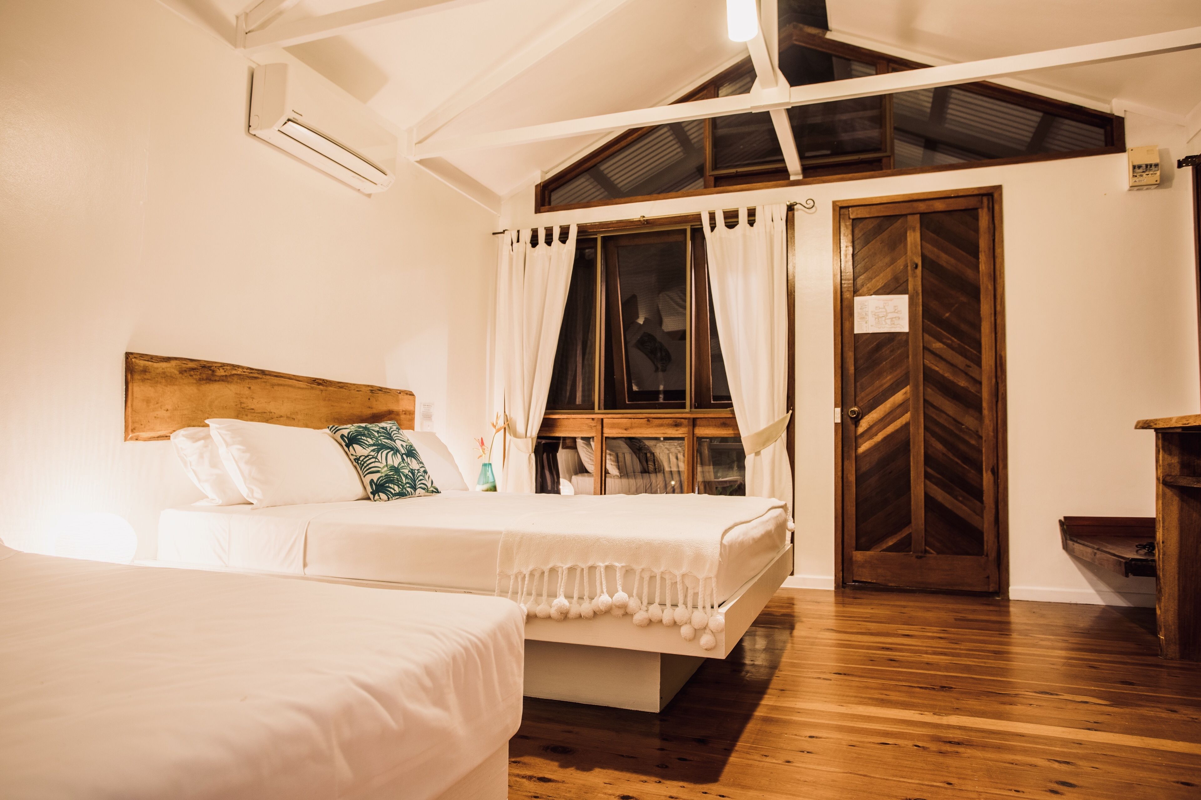Quarto com duas camas, soalho de madeira, uma janela grande com cortinas e uma porta de madeira.