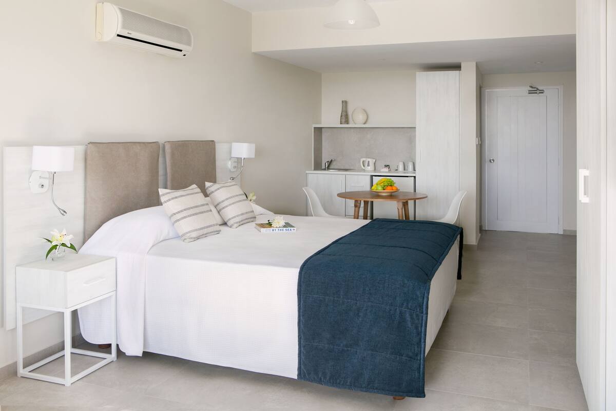 Quarto de hotel moderno com uma cama grande, mesas de cabeceira, uma pequena mesa redonda com uma tigela de frutas, um guarda-roupa branco e uma porta que dá para outro cômodo.