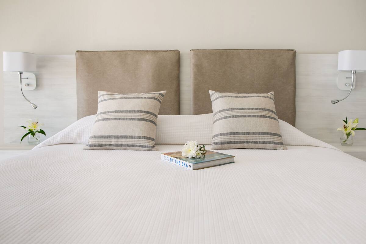 Cama bem arrumada com travesseiros listrados, um livro e um vaso com flores.