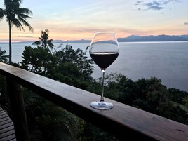 Um copo de vinho tinto num deck de madeira com vista para um corpo de água e montanhas ao longe.