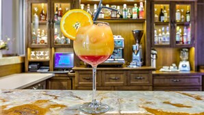 Um cocktail com uma fatia de laranja e um canudo, sobre um balcão de mármore num bar, com armários e estantes de garrafas ao fundo.