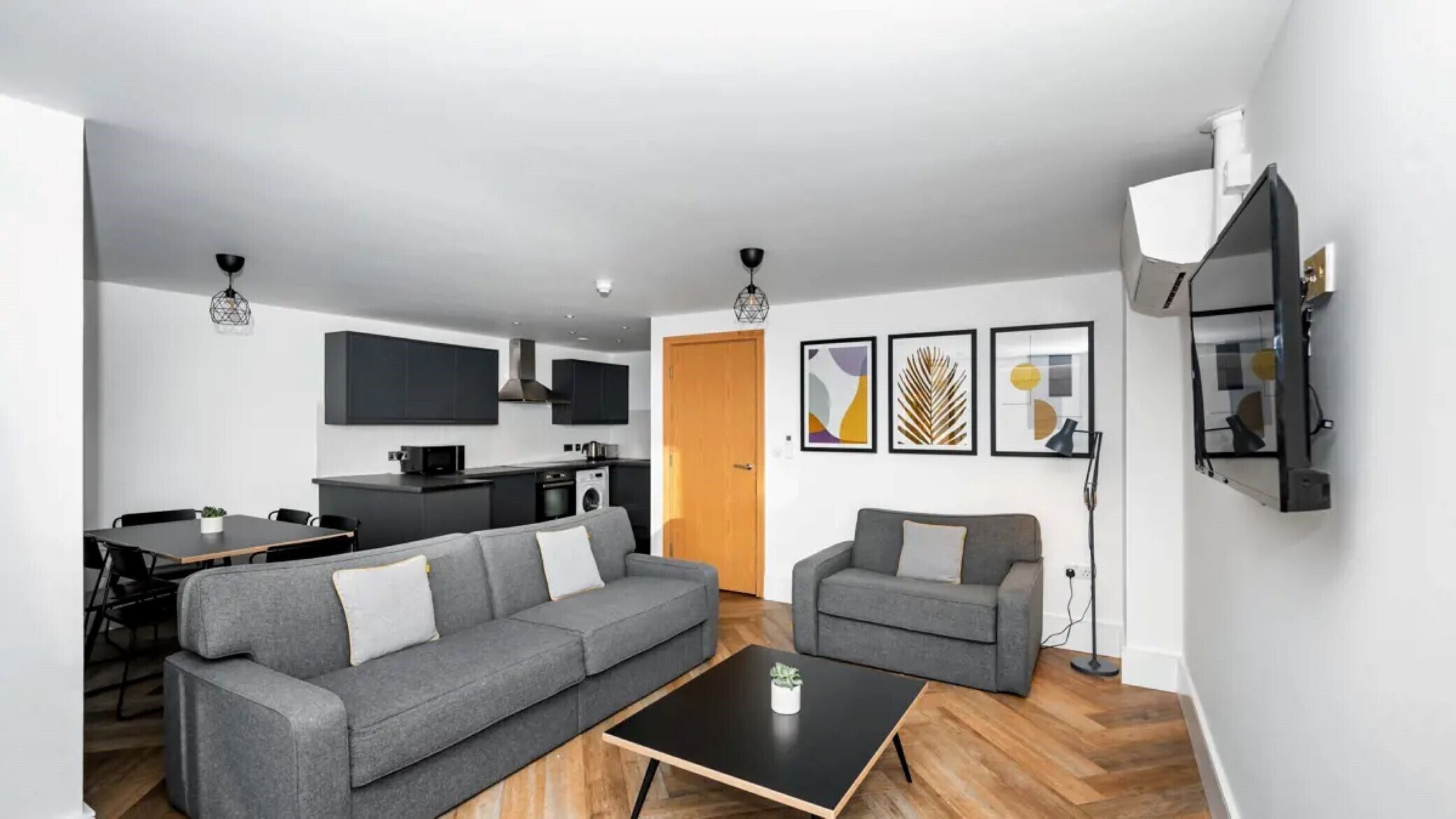 Sala de estar moderna com um sofá cinzento, uma poltrona cinzenta menor e uma mesa de centro preta. Há uma área de cozinha com armários escuros e um micro-ondas. O quarto tem piso de madeira e paredes brancas.
