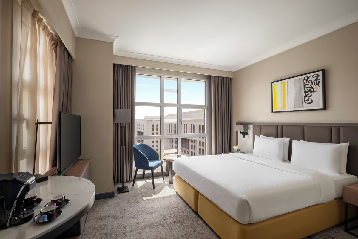 Quarto de hotel com uma cama grande, uma escrivaninha com cafeteira, uma cadeira e uma televisão.