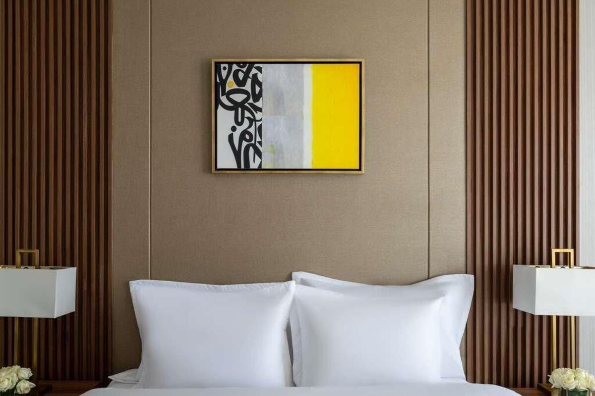 Quarto de hotel com cama, duas mesas de cabeceira, obra de arte na parede e painel de madeira.
