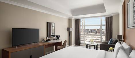 Quarto de hotel com uma cama grande, uma escrivaninha com televisão de tela plana, uma cadeira, uma mesinha e vista para a paisagem urbana através de janelas amplas.