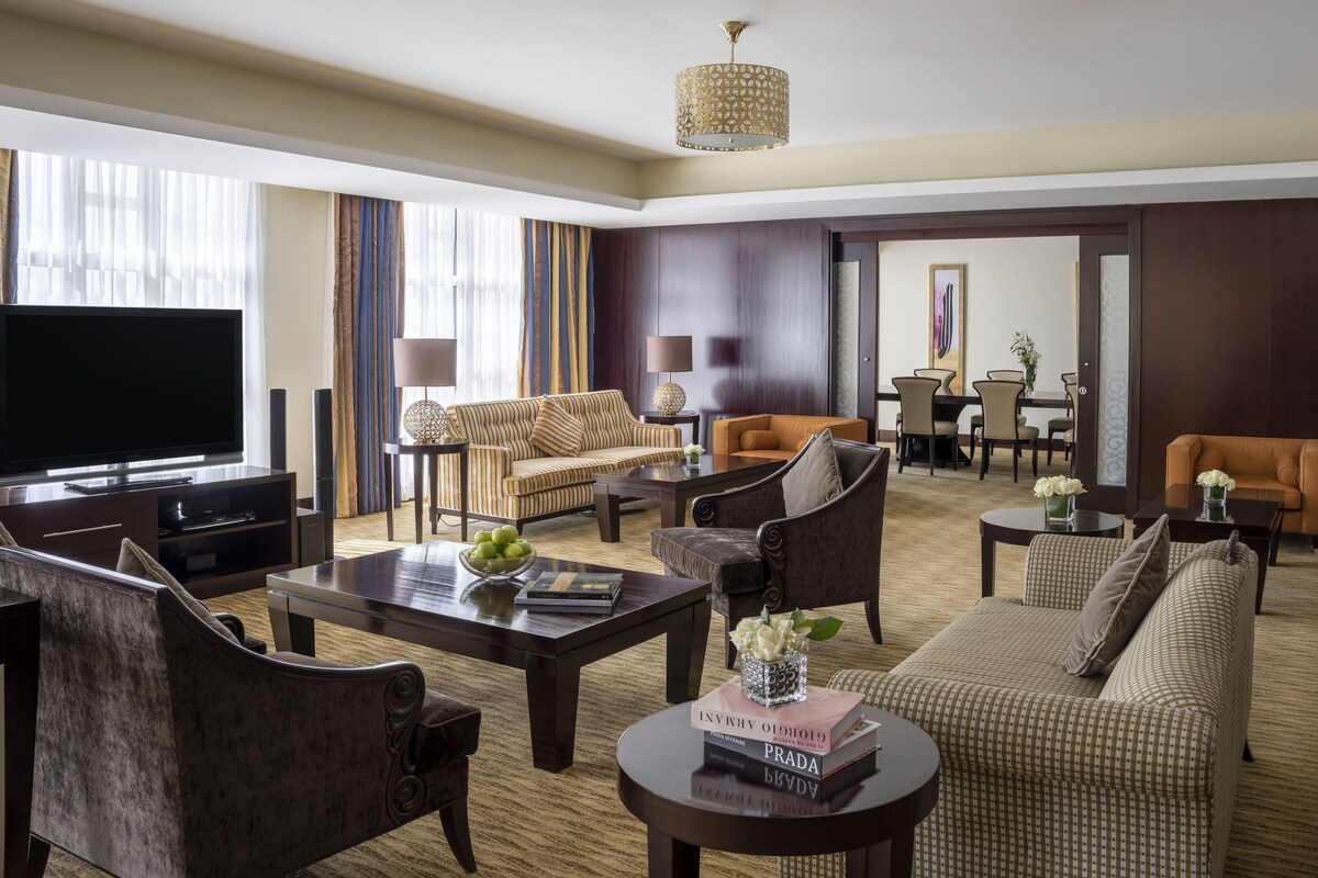 Uma sala de estar de hotel ampla, com uma televisão de tela grande, várias áreas de estar e uma mesa de jantar.