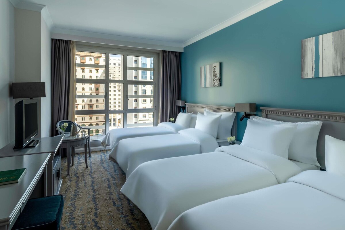 Quarto de hotel com duas camas, uma mesa de trabalho com televisão, uma cadeira e vista para edifícios através de janelas amplas.