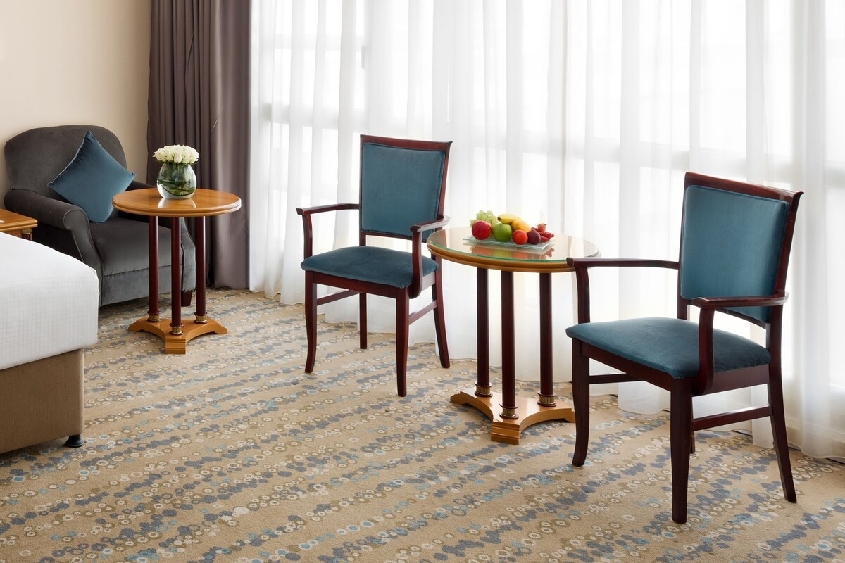 Quarto de hotel com cama, duas cadeiras, uma mesa pequena com frutas e um vaso de flores.