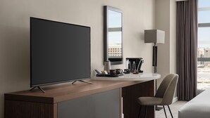 Quarto de hotel moderno com televisão de tela plana, escrivaninha com luminária, cadeira e uma janela grande com cortinas.