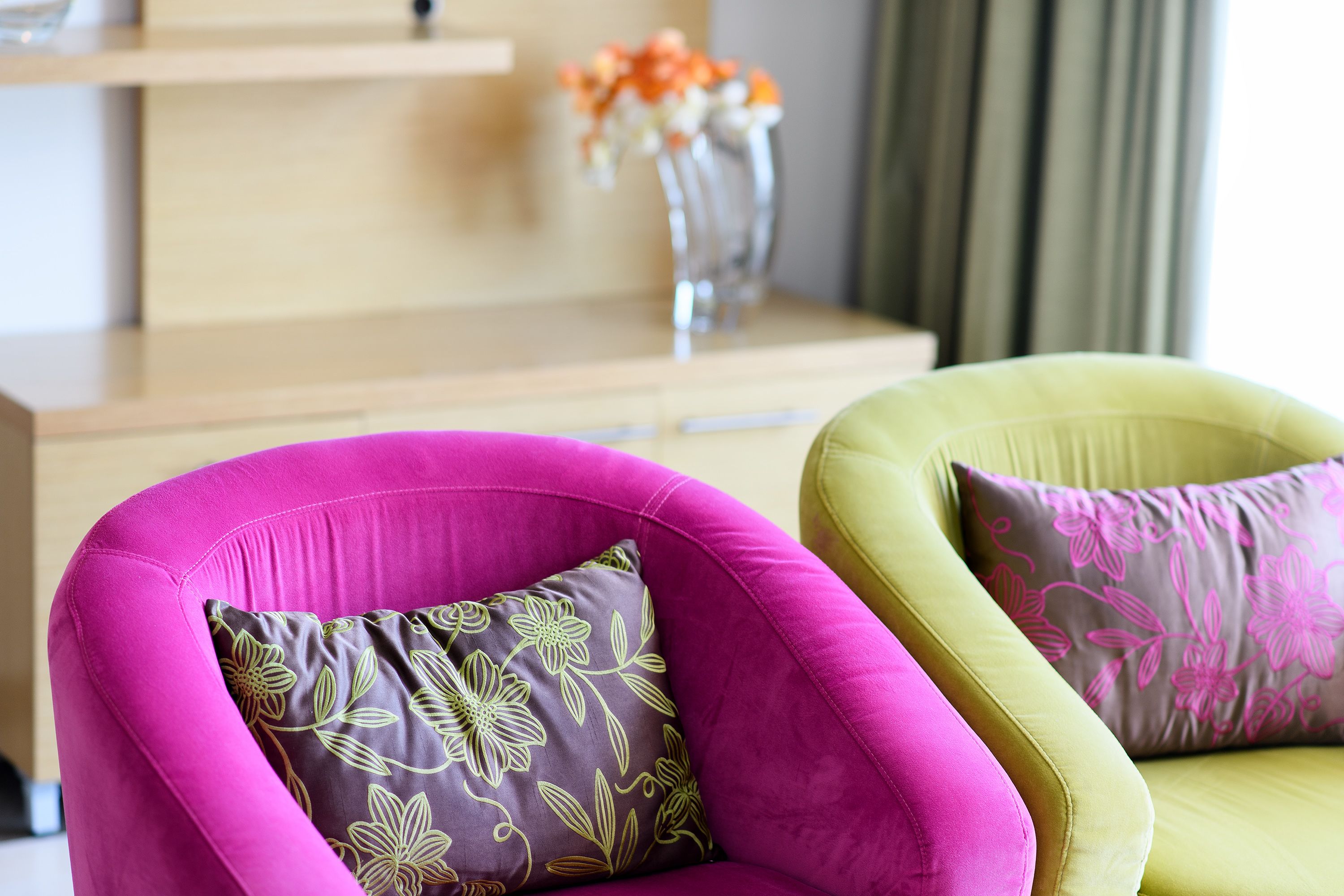 Duas poltronas com almofadas florais, uma mesa lateral de madeira e um vaso com flores ao fundo.