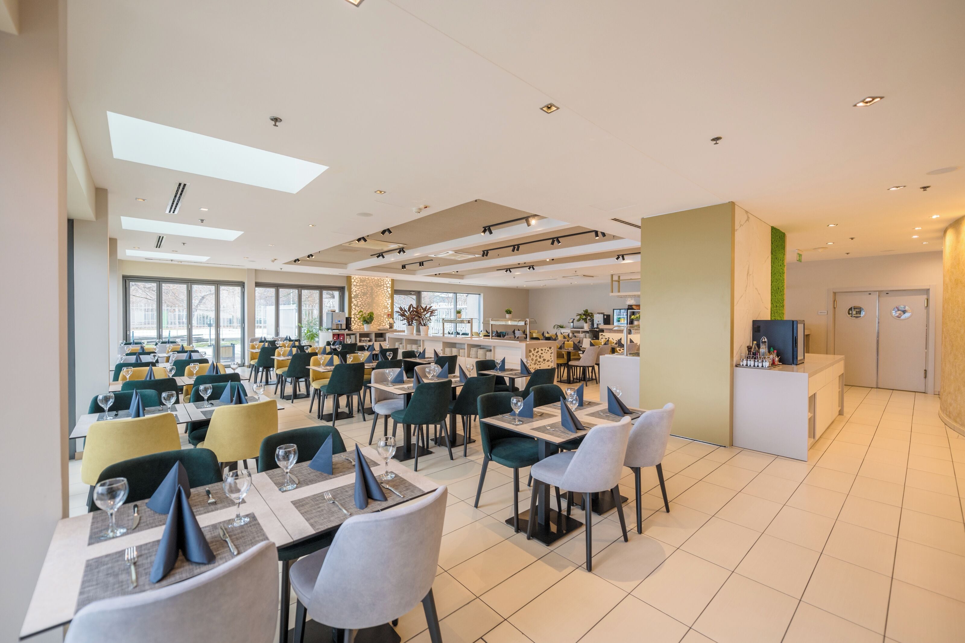 Uma sala de jantar espaçosa, com várias mesas e cadeiras, janelas amplas e um design moderno no teto.