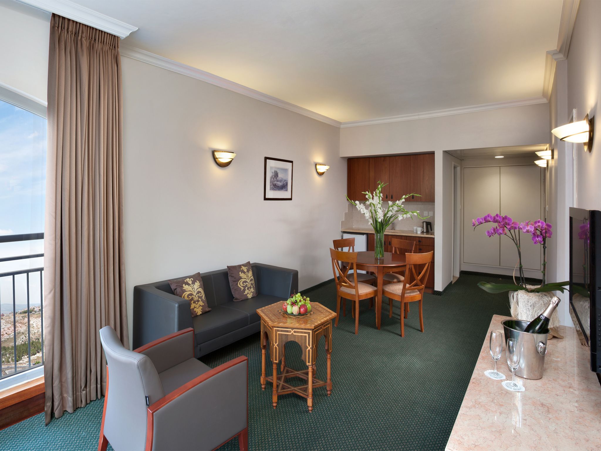 Quarto de hotel com sala de estar, mesa de jantar, cadeiras, uma pequena mesa com um vaso de flores, minibar e vista para o exterior.