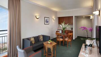 Quarto de hotel com sala de estar, mesa de jantar, cadeiras, uma pequena mesa com um vaso de flores, minibar e vista para o exterior.