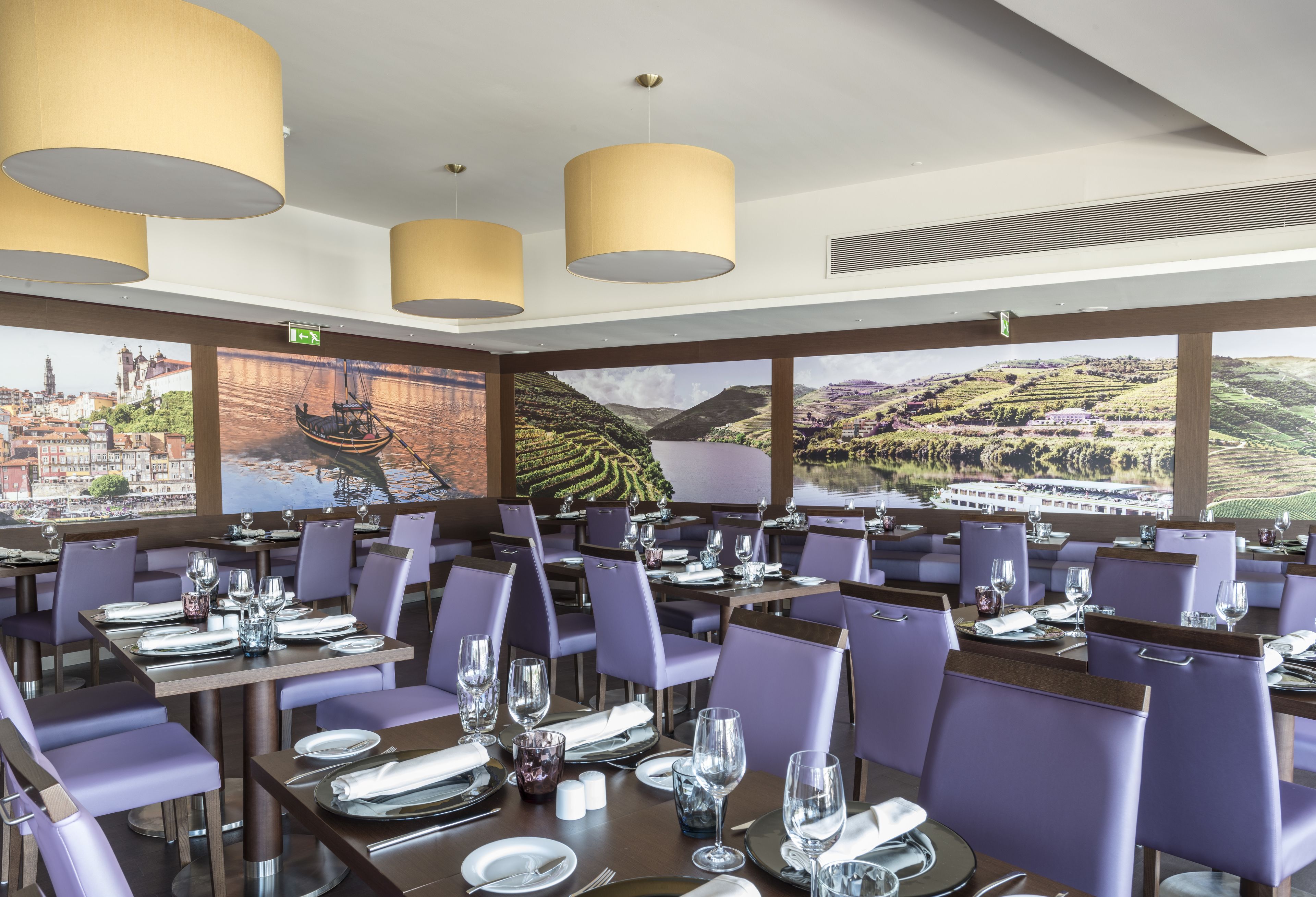 Restaurante com cadeiras e mesas roxas, grandes janelas com vistas panorâmicas e luminárias amarelas penduradas.