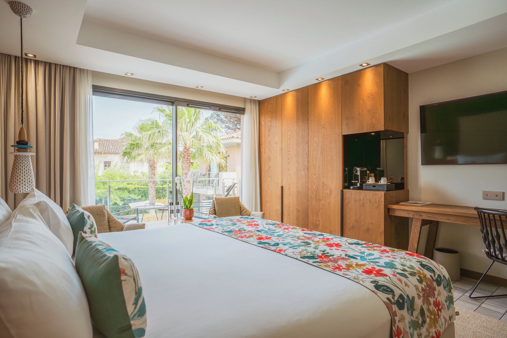 Quarto de hotel com uma cama grande, escrivaninha, televisão e varanda com vista para palmeiras.