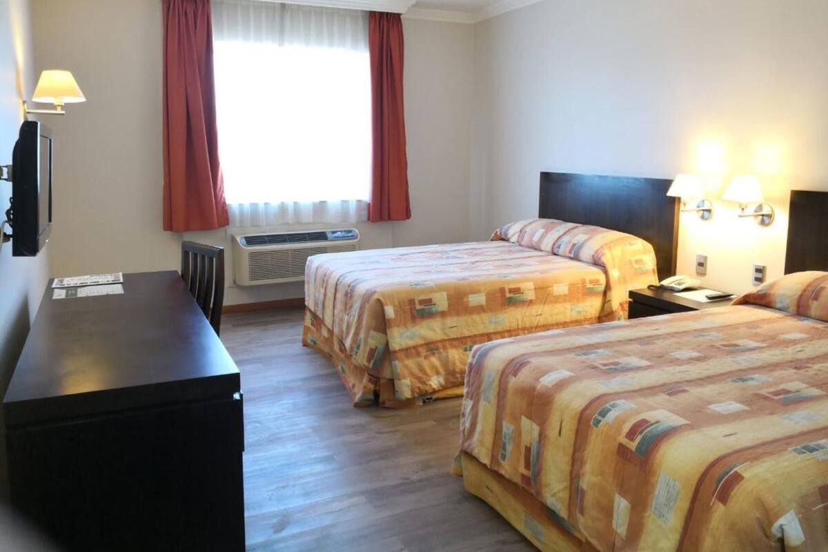 Quarto de hotel com duas camas, uma escrivaninha, uma cadeira, um abajur e janela com cortinas.