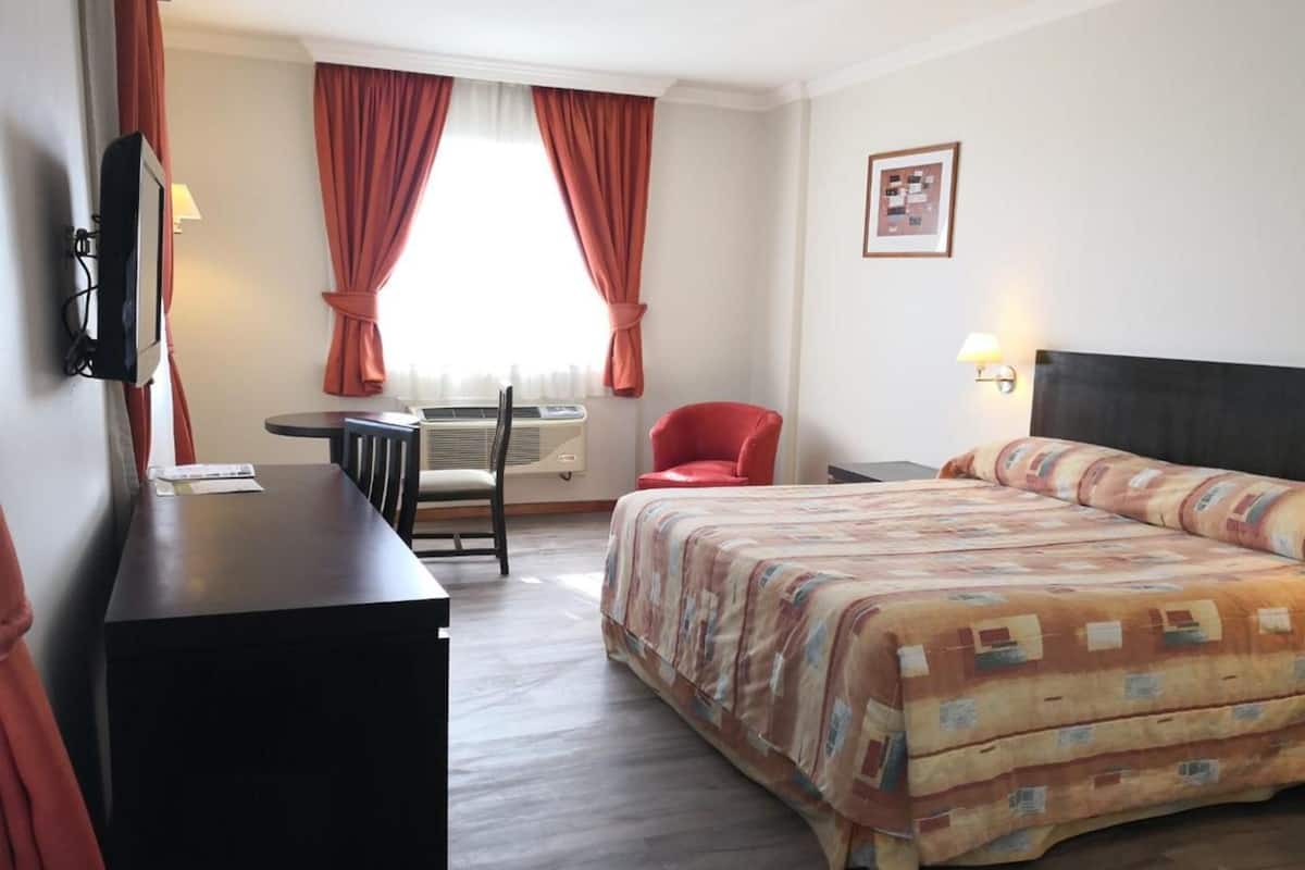 Quarto de hotel com cama, escrivaninha, cadeira e televisão.