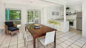 Uma cozinha moderna com armários brancos, uma geladeira de aço inoxidável e uma área de refeições com mesa de madeira e cadeiras brancas.