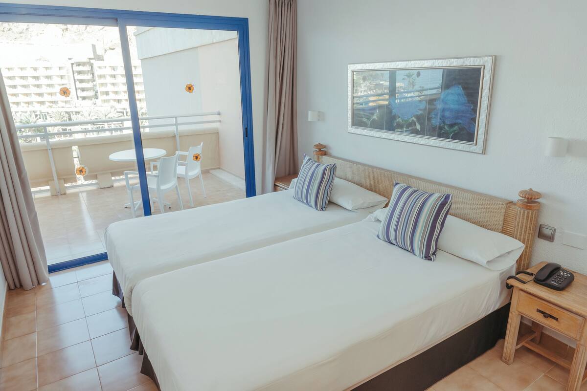 Quarto de hotel com duas camas, varanda com mesa e cadeiras, e vista para a cidade.