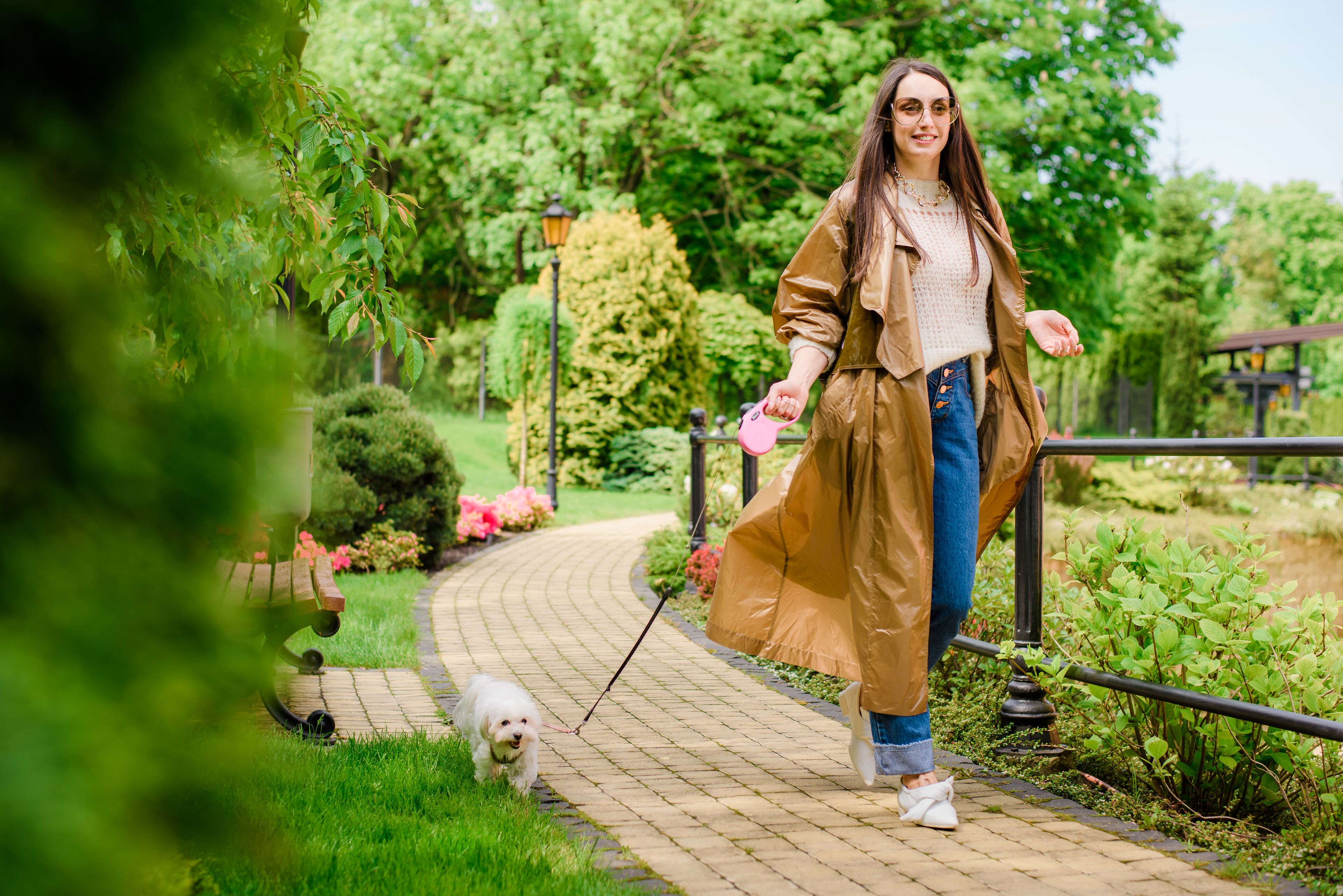 Uma pessoa passeando com um cachorro em um caminho pavimentado em um parque, com vegetação e um poste de luz.