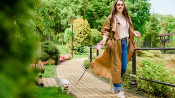 Uma pessoa passeando com um cachorro em um caminho pavimentado em um parque, com vegetação e um poste de luz.