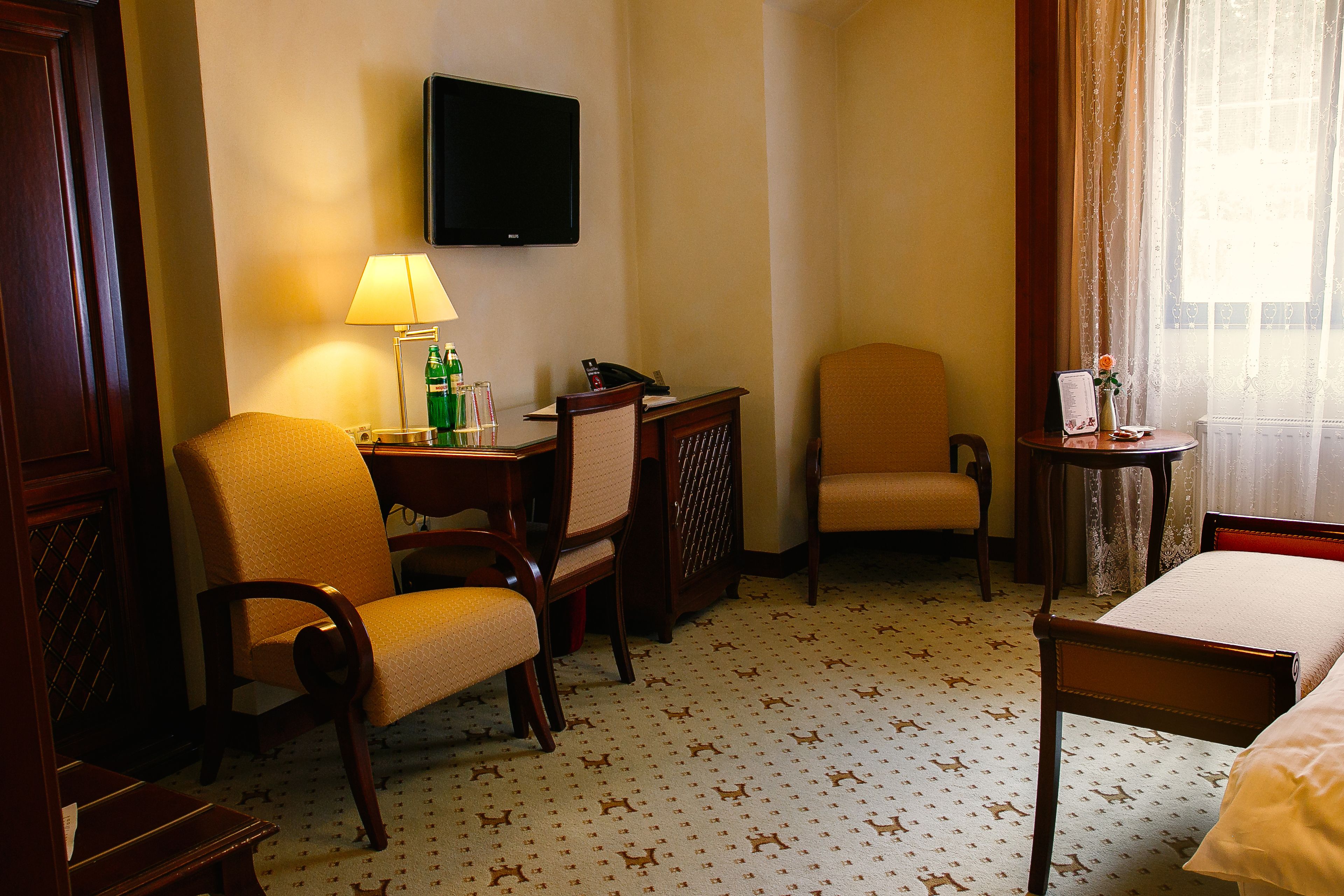 Quarto de hotel com cama, duas cadeiras, escrivaninha com luminária, televisão e janela com cortinas.