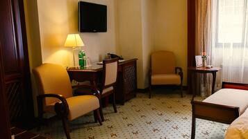 Quarto de hotel com cama, duas cadeiras, escrivaninha com luminária, televisão e janela com cortinas.