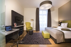 Quarto de hotel moderno com escrivaninha, laptop, poltrona verde e cama com cabeceira verde.