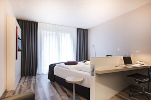 Quarto de hotel moderno com cama, escrivaninha, cadeira e uma janela grande com cortinas.