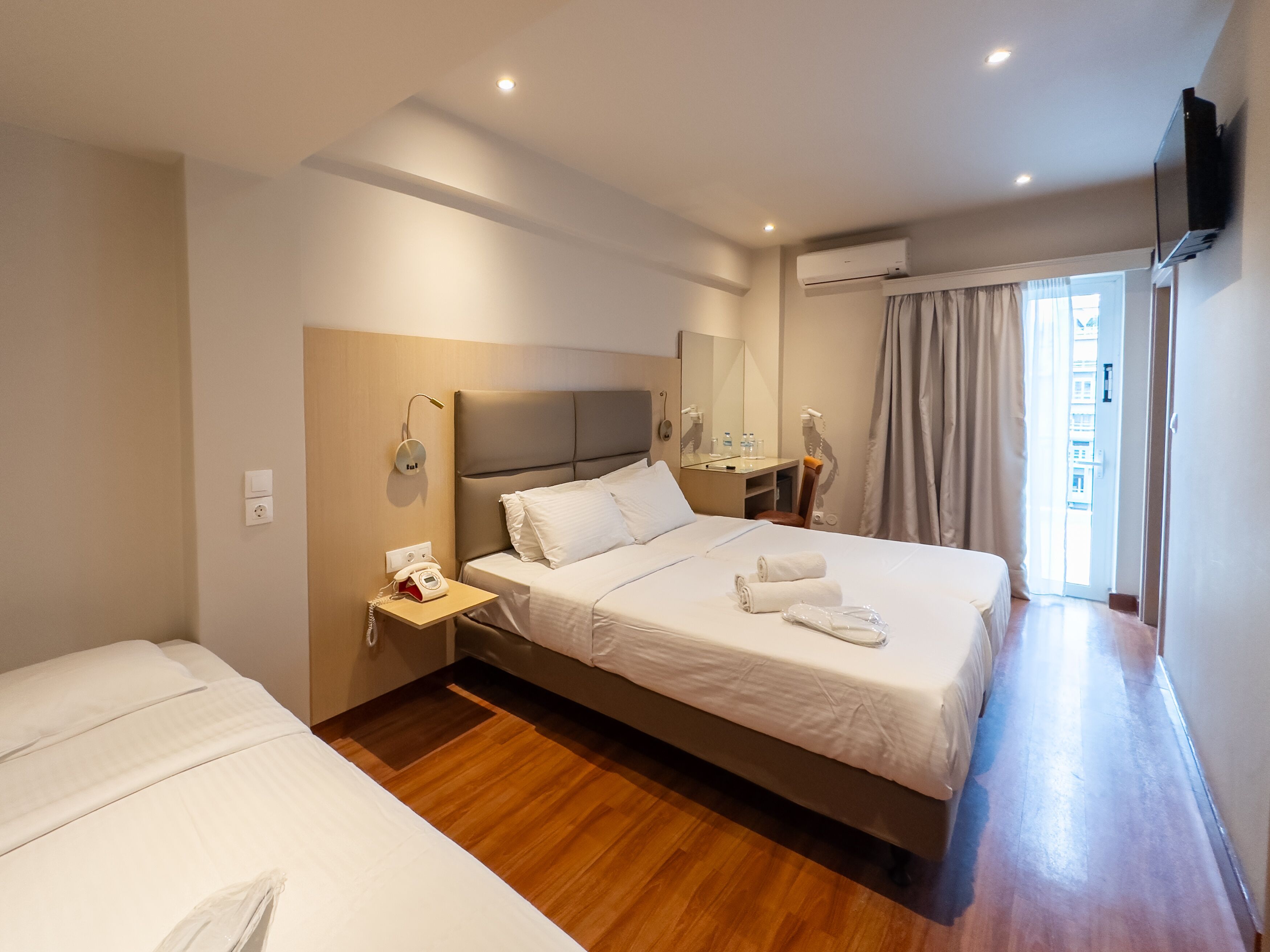 Quarto de hotel com uma cama grande, uma cama menor, mesa de cabeceira com telefone, espelho e ar condicionado.