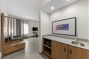Quarto de hotel moderno com guarda-roupa de madeira, televisão de tela plana, quadro com placa da Texas State Railroad e área de cozinha com pia e micro-ondas.
