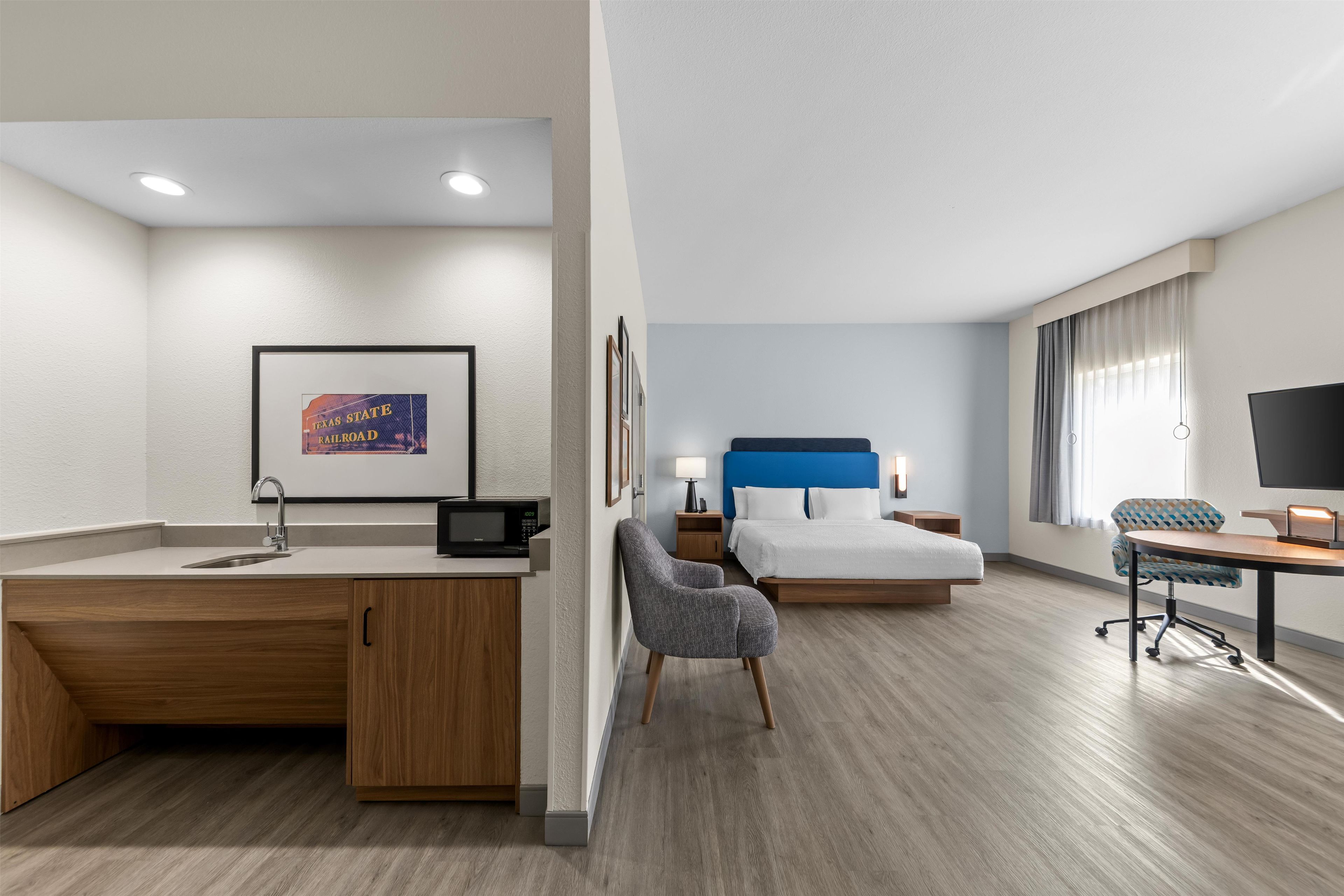Quarto de hotel com cama, escrivaninha, cadeira, televisão e um quadro na parede.