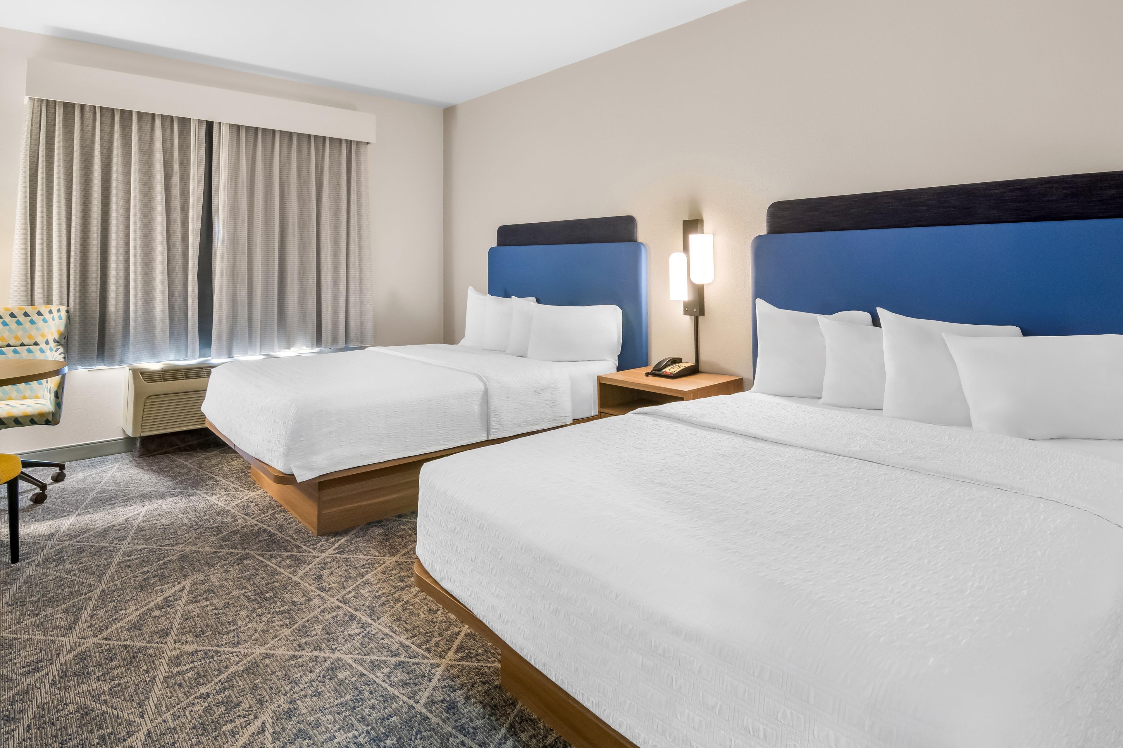 Quarto de hotel com duas camas, uma escrivaninha, uma cadeira, um abajur e uma janela com cortinas.