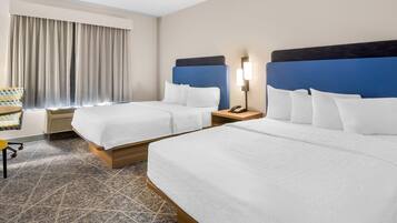 Quarto de hotel com duas camas, uma escrivaninha, uma cadeira, um abajur e uma janela com cortinas.