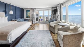 Quarto de hotel moderno com uma cama grande, um sofá, uma poltrona, uma mesa pequena e vista para o mar através de amplas janelas.