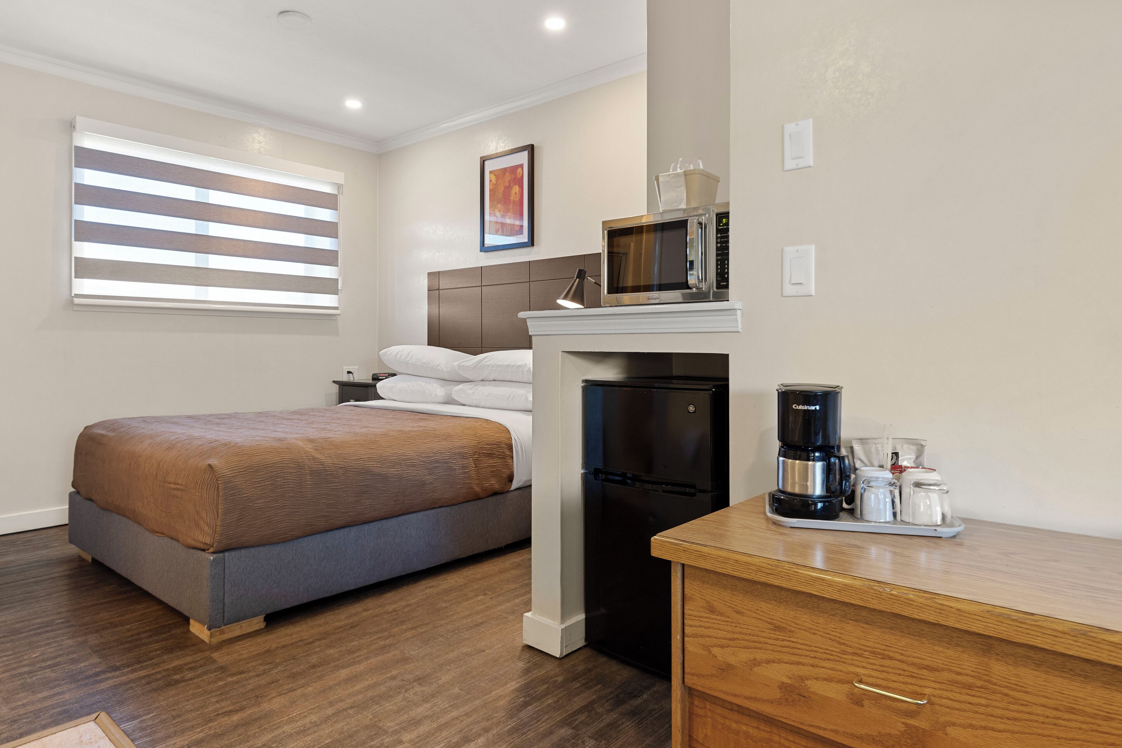 Quarto com cama, geladeira, micro-ondas e cafeteira sobre uma mesa de madeira.