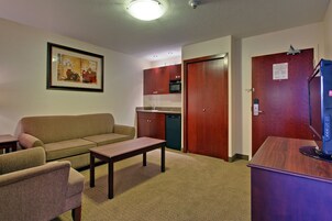 Uma sala de estar compacta com um sofá, uma mesinha e um espaço de cozinha americana com armários de madeira e micro-ondas.