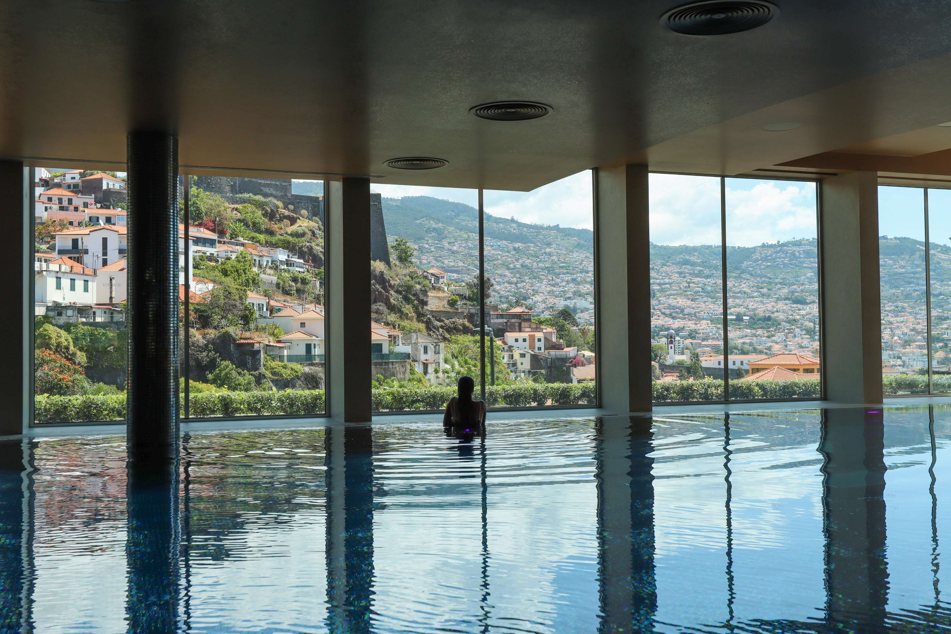 Uma grande piscina interior com vista para uma cidade situada numa encosta, através de amplas janelas.
