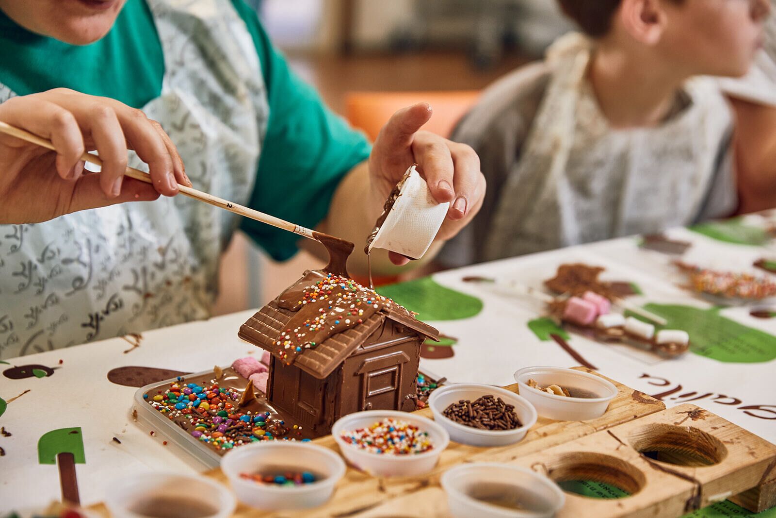 Uma pessoa derretendo chocolate sobre um modelo de chocolate em formato de casa, com confeitos, rodeada por recipientes com confeitos e outras decorações de chocolate.