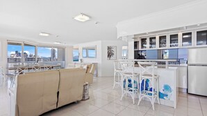 Uma sala de estar moderna com um sofá bege, cadeiras brancas e uma área de cozinha com geladeira e armários.