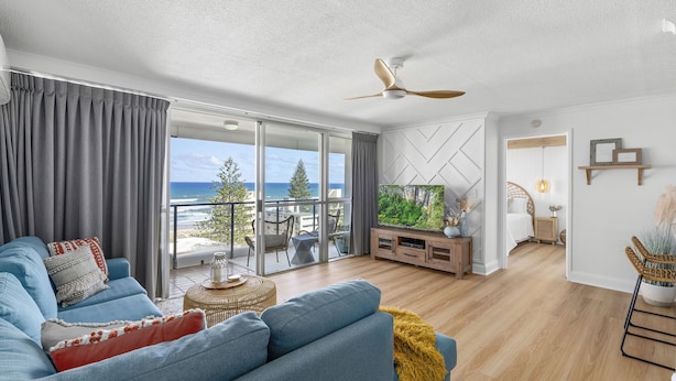 Uma sala de estar moderna com um sofá azul, um ventilador de teto, uma janela grande com vista para o oceano e piso de madeira.
