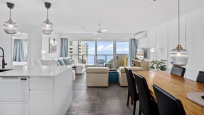 Uma sala de estar moderna com uma mesa de jantar de madeira, sofás brancos e uma área de cozinha com bancada branca e lava-louças.