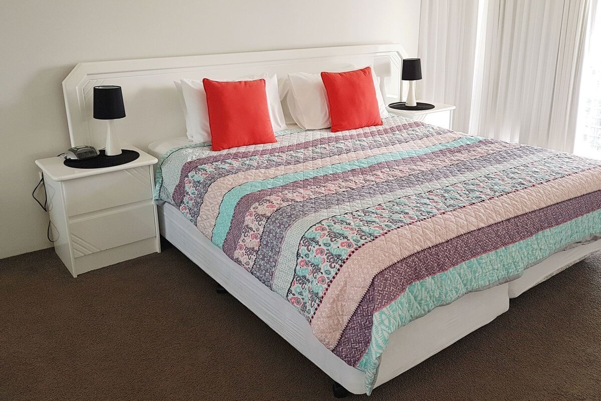 Uma cama bem arrumada com um edredom estampado, duas mesas de cabeceira brancas com abajures e uma janela com cortinas.