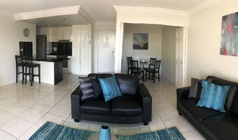 Uma sala de estar moderna com um sofá de couro preto, uma área de jantar com uma mesa pequena e cadeiras, e uma cozinha com armários brancos e bancada preta.