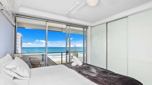 Quarto de hotel com uma cama grande, vista para o oceano, ventilador de teto e porta de vidro deslizante.