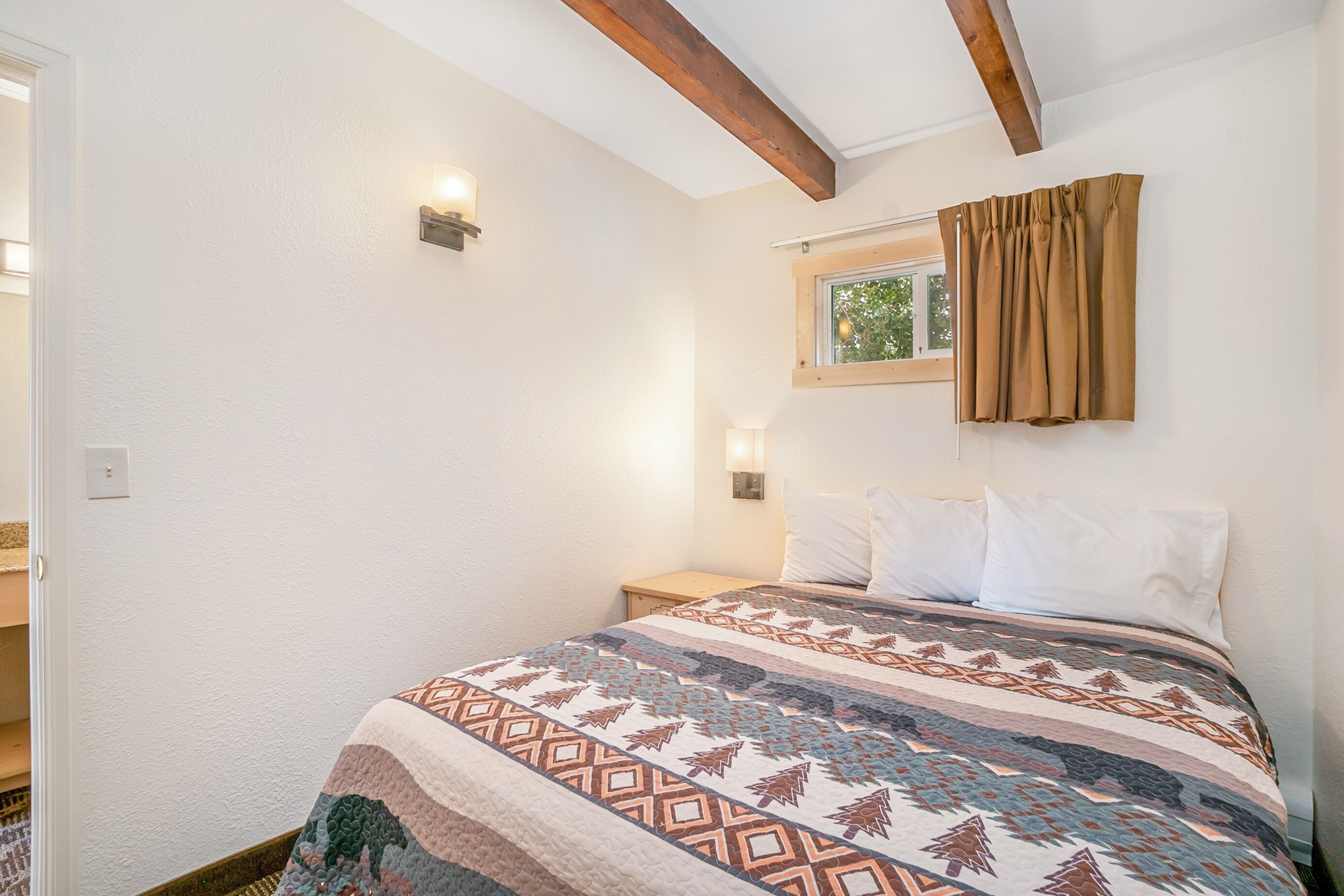 Quarto com cama, janela com cortinas, mesa de cabeceira e luminária fixada na parede.