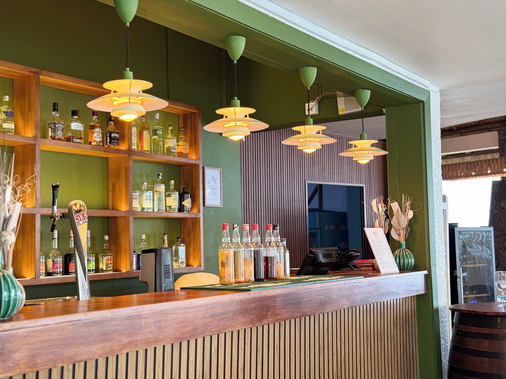 Uma área de bar com prateleiras de madeira repletas de garrafas de bebidas, balcão e luminárias pendentes.