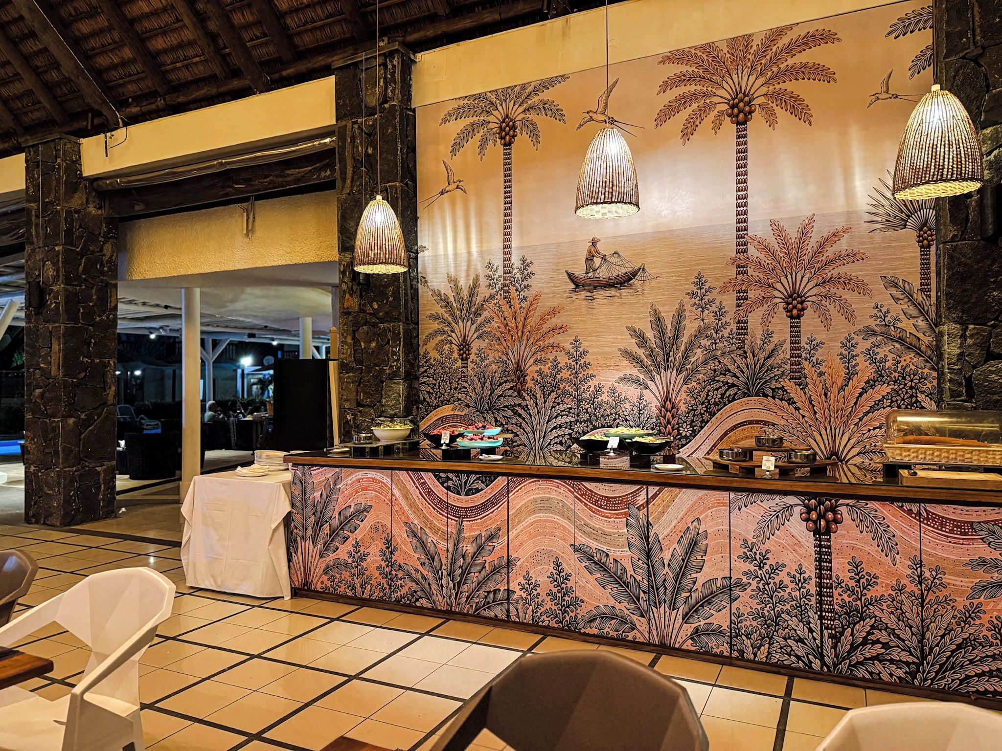 Uma área de recepção com parede decorativa mostrando palmeiras e um pescador, balcão com luminária de palha e piso de azulejos.